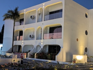 Um edifício moderno de vários andares, com varandas, iluminação externa e aviso de chão molhado.