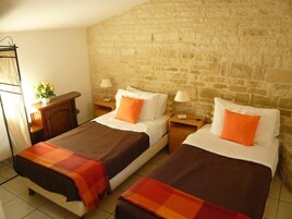 Quarto com duas camas, cada uma com cobertor e almofadas, uma mesinha de cabeceira de madeira, um abajur e uma pequena mesa com uma planta.