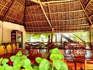Um restaurante com telhado de palha, mesas e cadeiras de madeira, e um mural na parede.