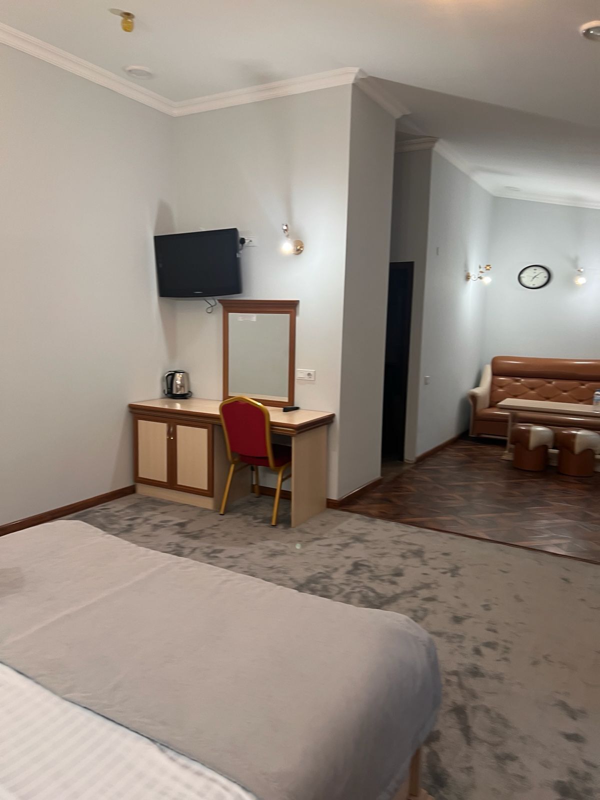 Quarto de hotel com uma cama, escrivaninha com cadeira, televisão, espelho e área de estar com sofá e poltronas.