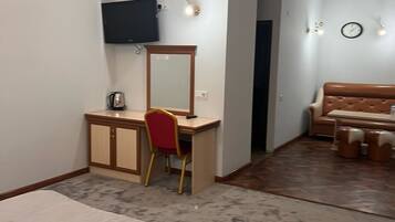 Quarto de hotel com uma cama, escrivaninha com cadeira, televisão, espelho e área de estar com sofá e poltronas.