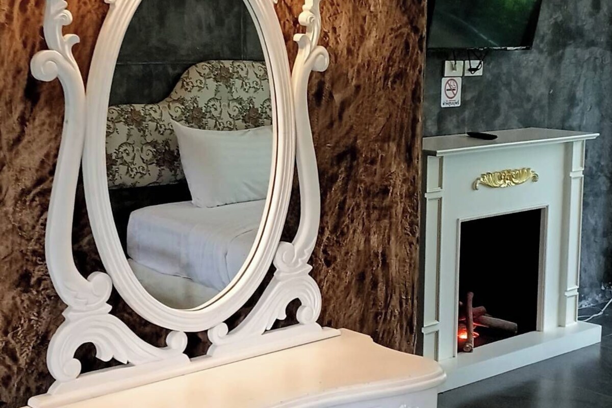 Uma toucador branca com espelho decorado, cama com roupa de cama estampada e lareira com televisão em cima.