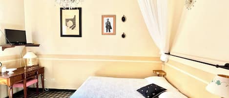 Quarto de hotel com cama, escrivaninha com abajur, televisão em uma estante e lustre.