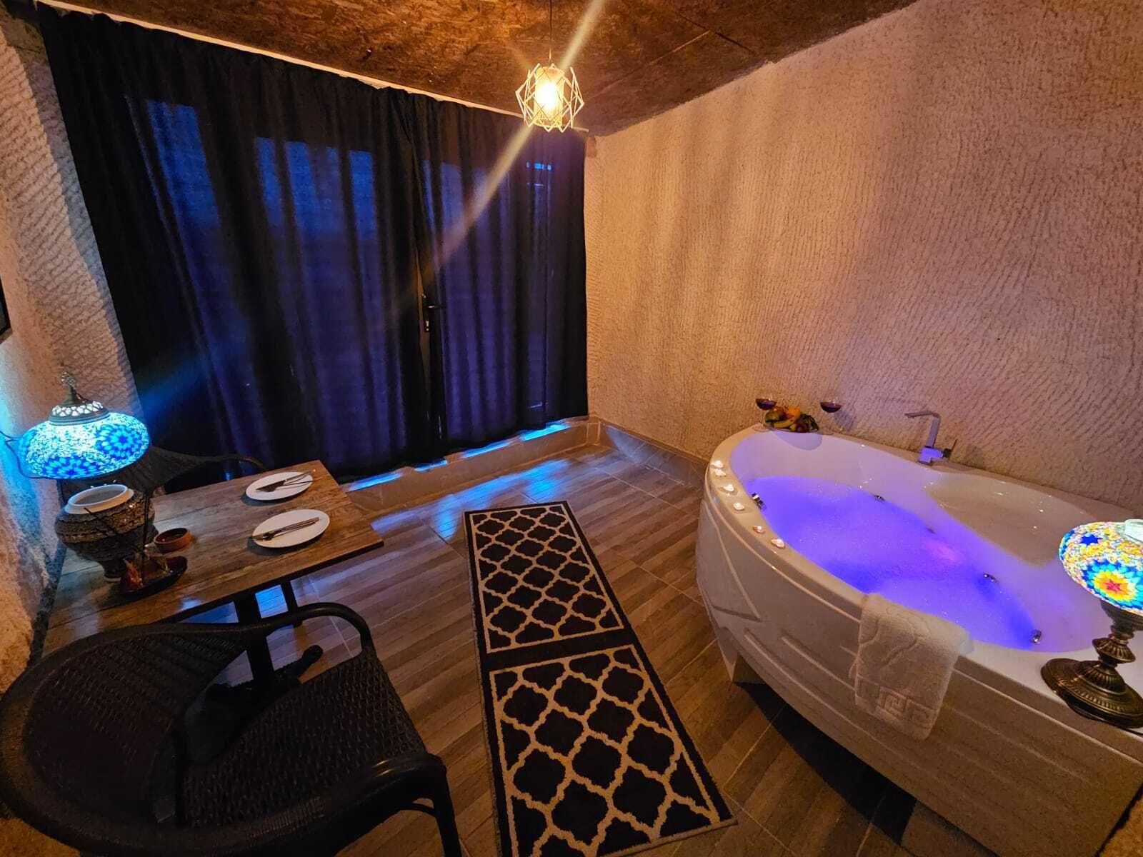 Quarto com piso de madeira, uma cadeira preta, uma mesinha com pratos e uma luminária, uma banheira com velas e uma janela com cortinas.