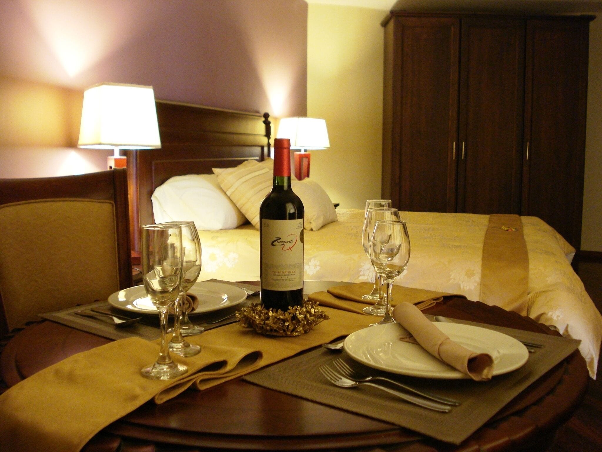 Uma mesa de jantar bem posta com uma garrafa de vinho, copos e talheres, uma cama com almofadas, e um guarda-roupa de madeira num quarto de hotel.