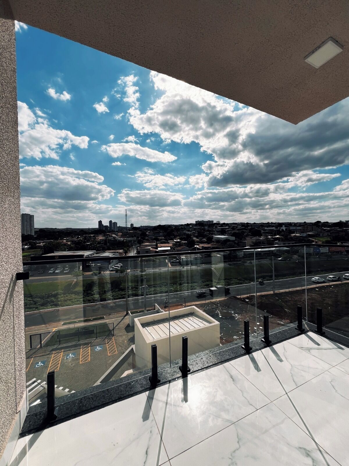 Um edifício alto com varanda de vidro, vista para a cidade e um estacionamento.