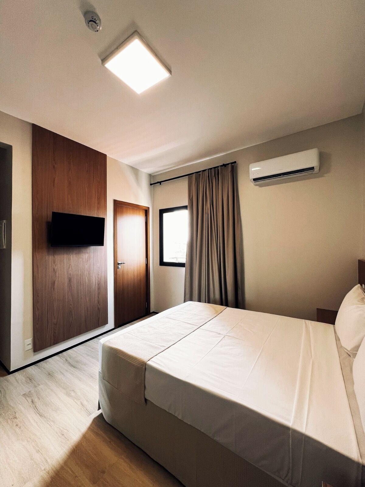 Quarto de hotel com cama, guarda-roupa, televisão, ar condicionado e janela com cortinas.