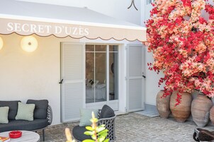 Exterior de hotel com uma placa indicando 'SECRET D'UZES', área coberta para estar ao ar livre com um sofá e almofadas, e grandes vasos de flores.