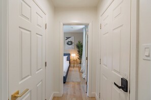 Um corredor leva a um quarto com uma cama, mesa de cabeceira, luminária e planta em vaso.