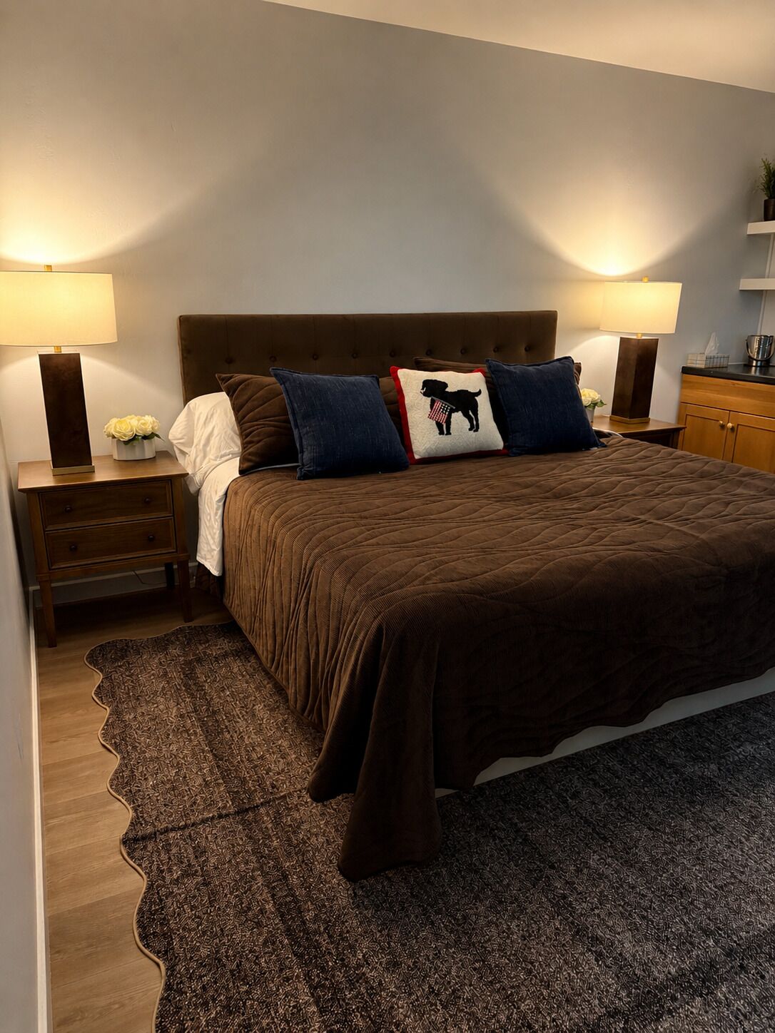 Uma cama bem arrumada com um edredom castanho, duas luminárias de cabeceira, uma mesa de cabeceira com um vaso de flores e um tapete estampado.