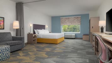Quarto de hotel com cama, escrivaninha, cadeira, televisão e janela com vista para árvores.