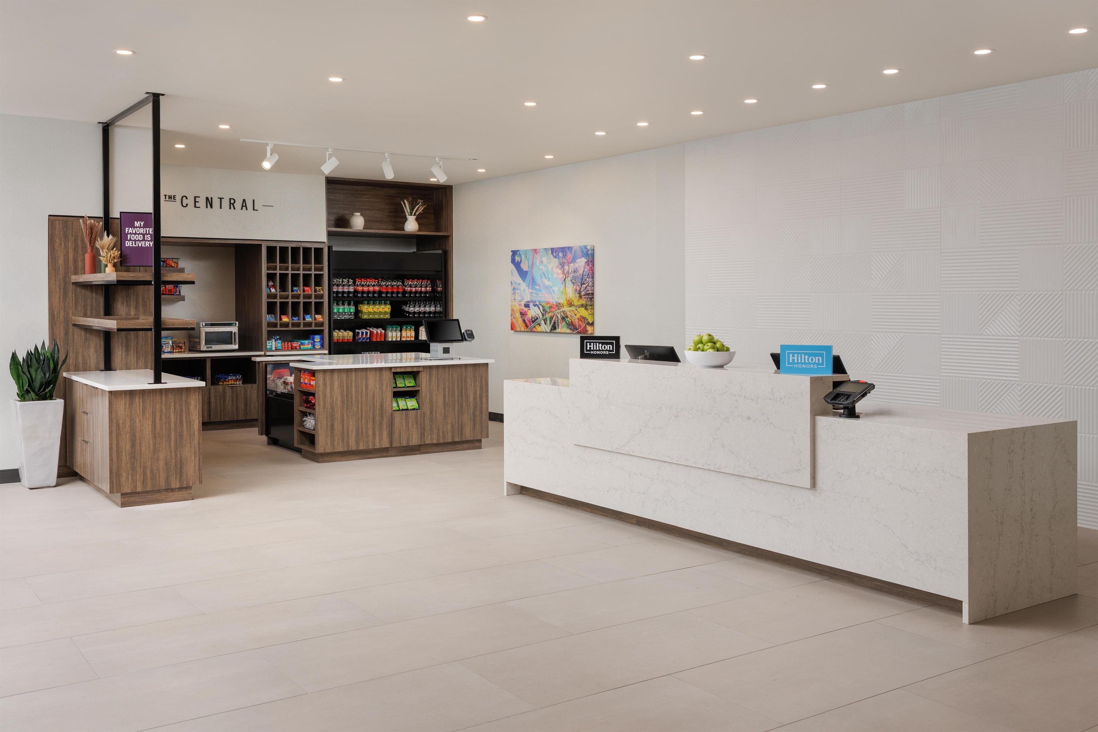 Uma área de recepção de hotel com design moderno, incluindo um balcão de atendimento, um quiosque de autoatendimento e um balcão de entrega de comida.