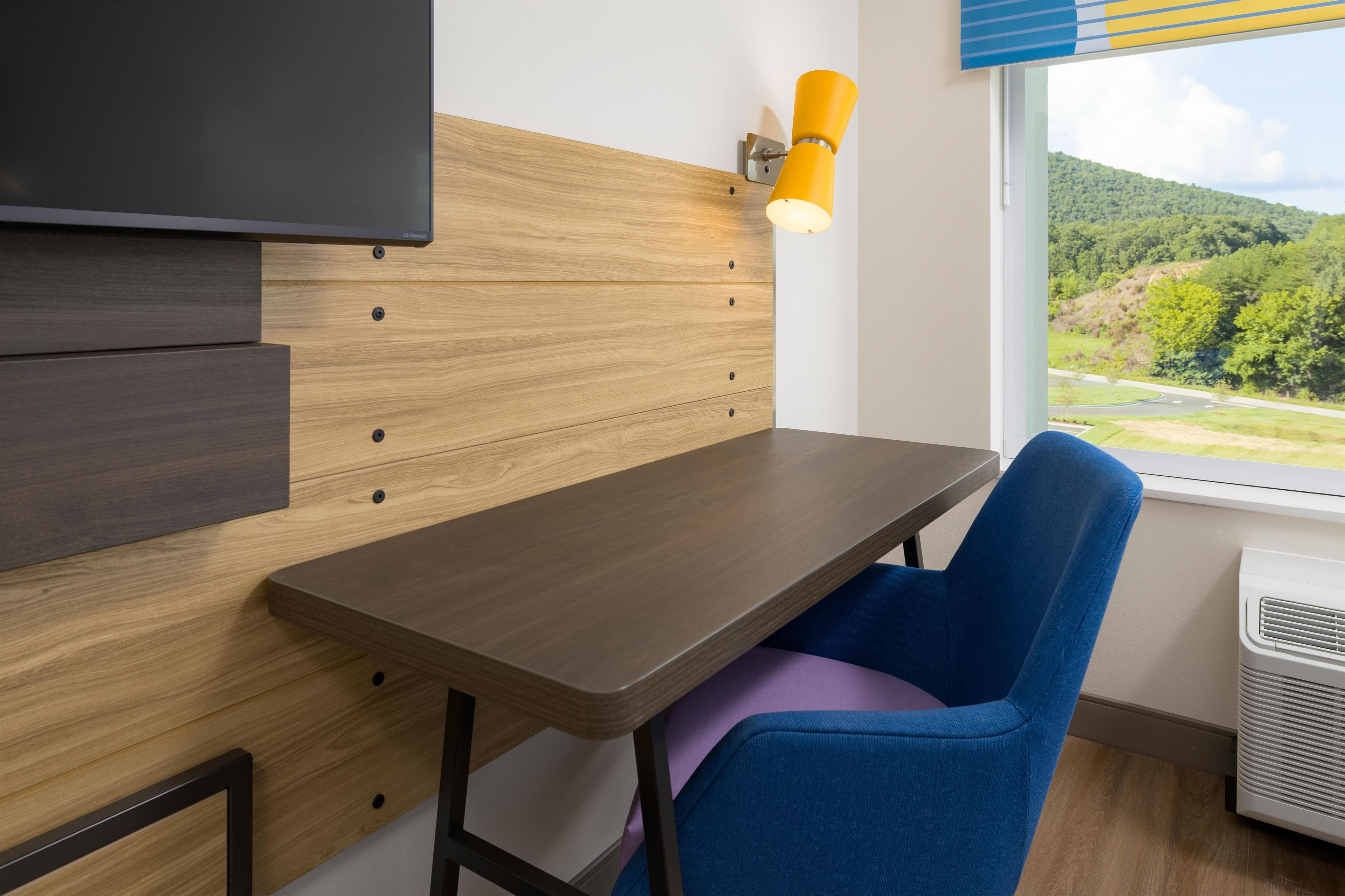 Quarto moderno com painel de parede de madeira, uma mesa, uma cadeira azul e janela com vista para uma paisagem verde.
