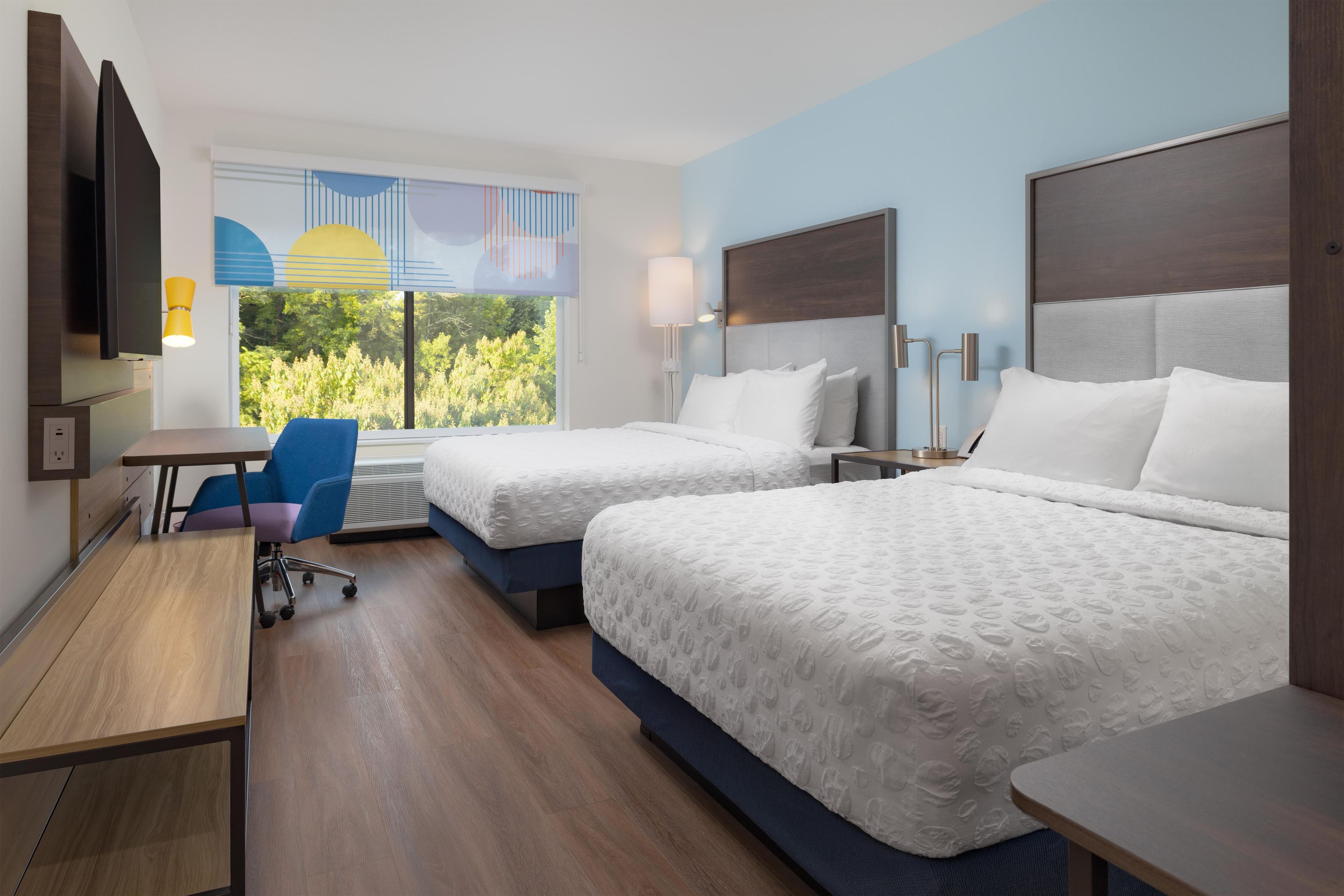 Quarto de hotel com duas camas, uma escrivaninha, uma cadeira e janela com vista para árvores.