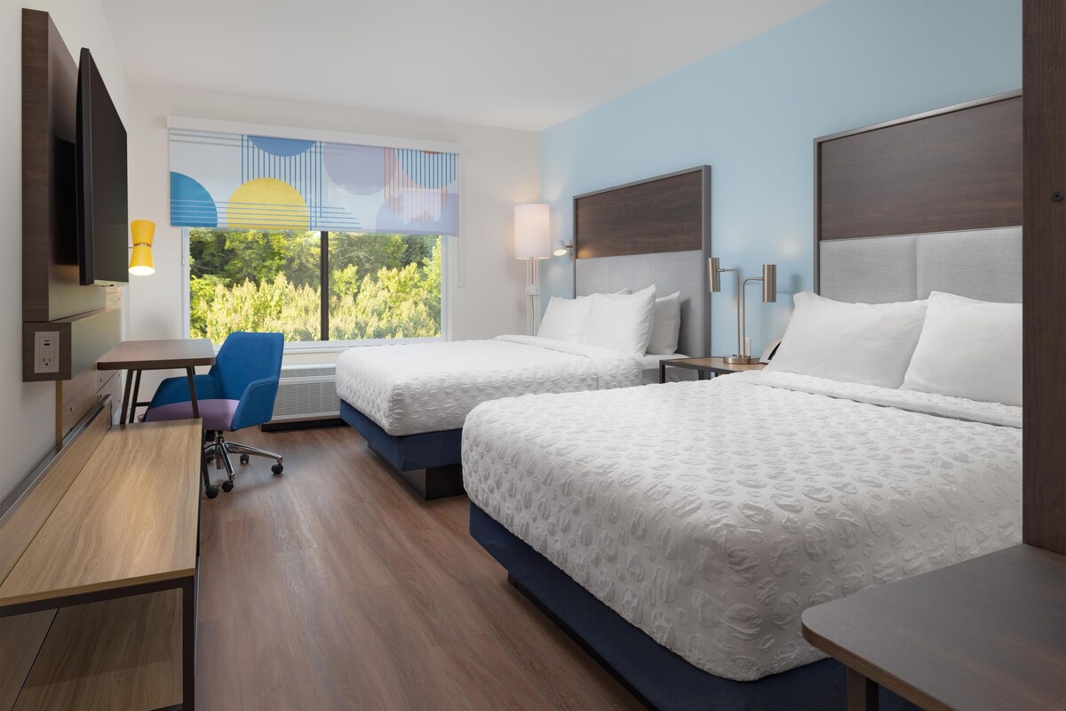 Quarto de hotel com duas camas, uma escrivaninha, uma cadeira e janela com vista para árvores.