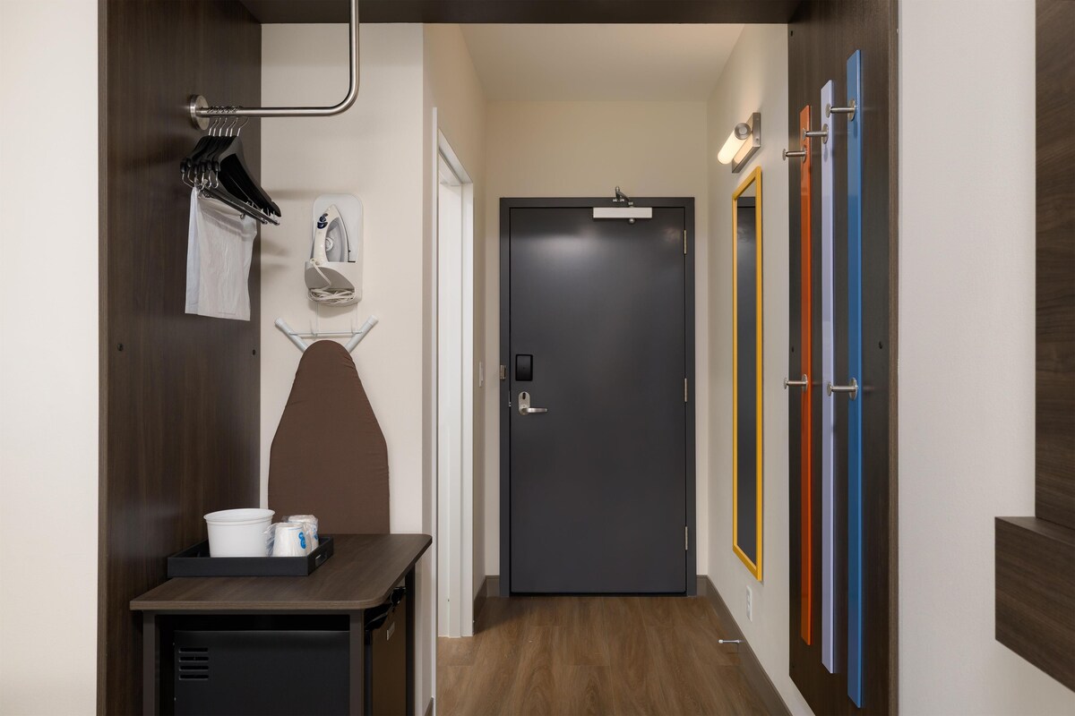 Um corredor com uma porta escura, um porta-roupas com cabides, uma pequena mesa com um cesto e um painel de passar roupa fixado na parede.