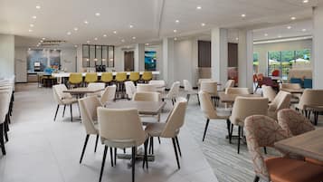 Uma sala de jantar ampla com várias mesas e cadeiras, uma cozinha ao fundo e vista para o exterior.