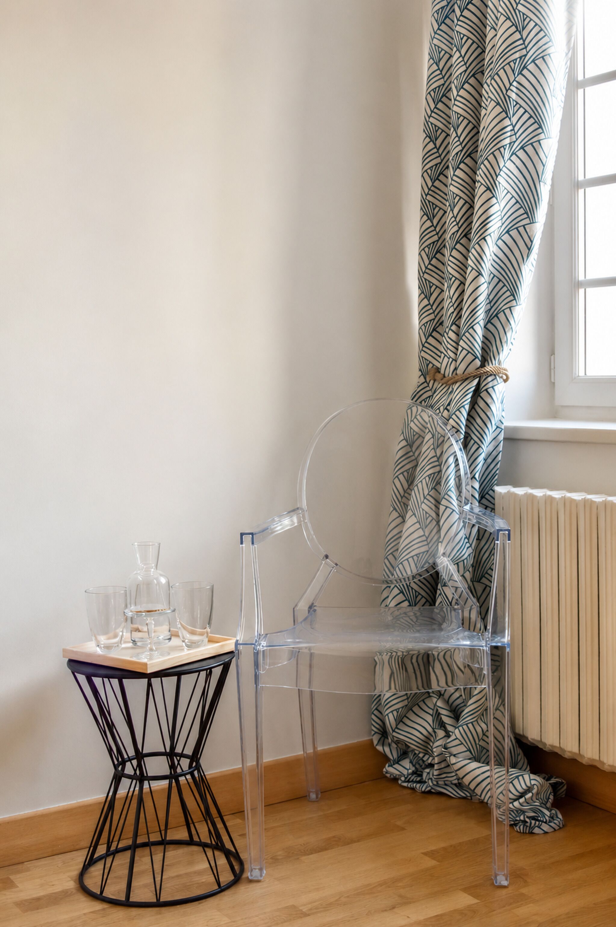 Uma mesa de vidro transparente com base de arame preto, uma cadeira transparente e uma cortina estampada.