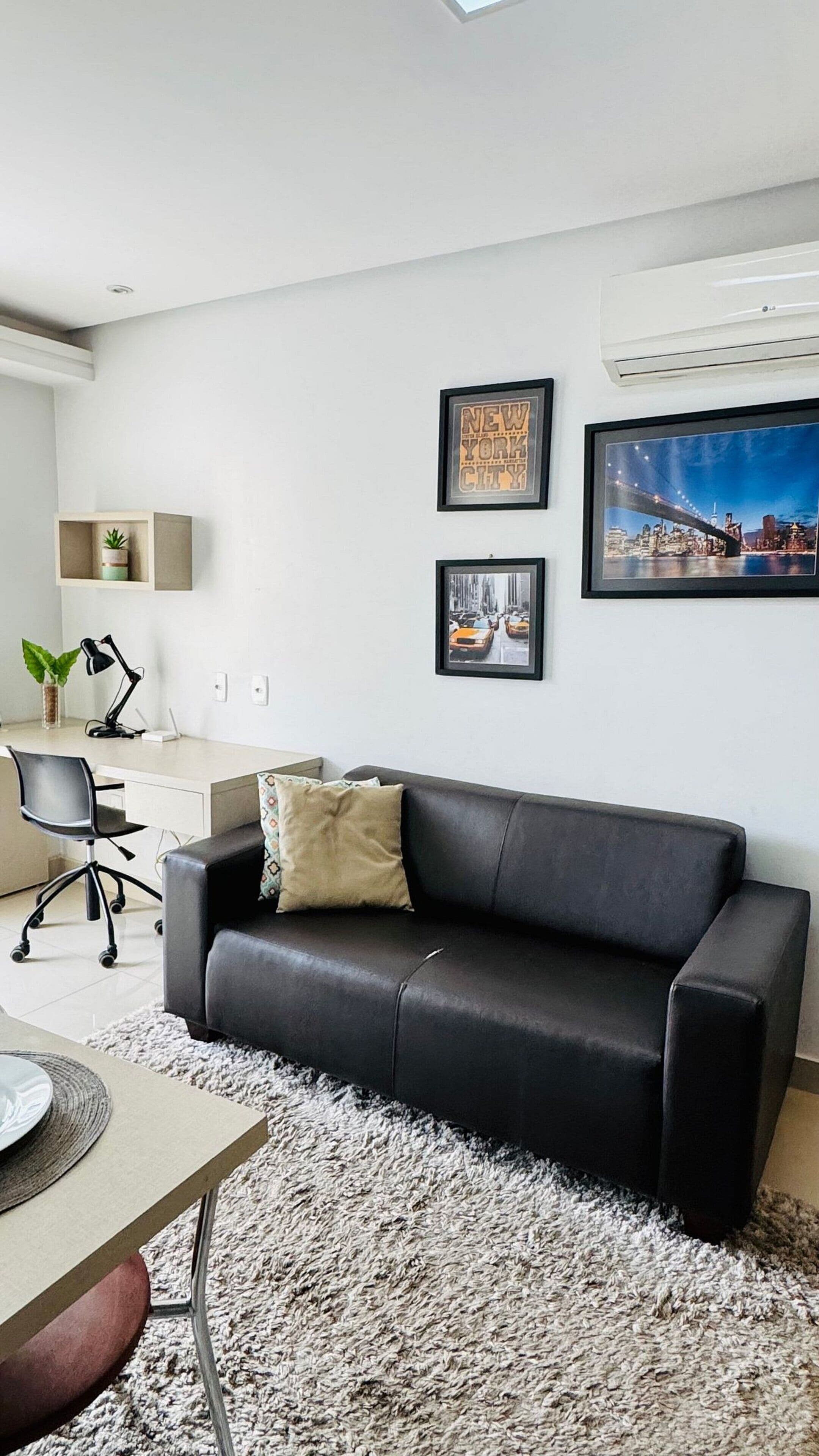 Uma sala de estar moderna com um sofá de couro preto, uma escrivaninha com um computador, uma cadeira e uma pequena planta. Há quadros nas paredes e uma janela grande.