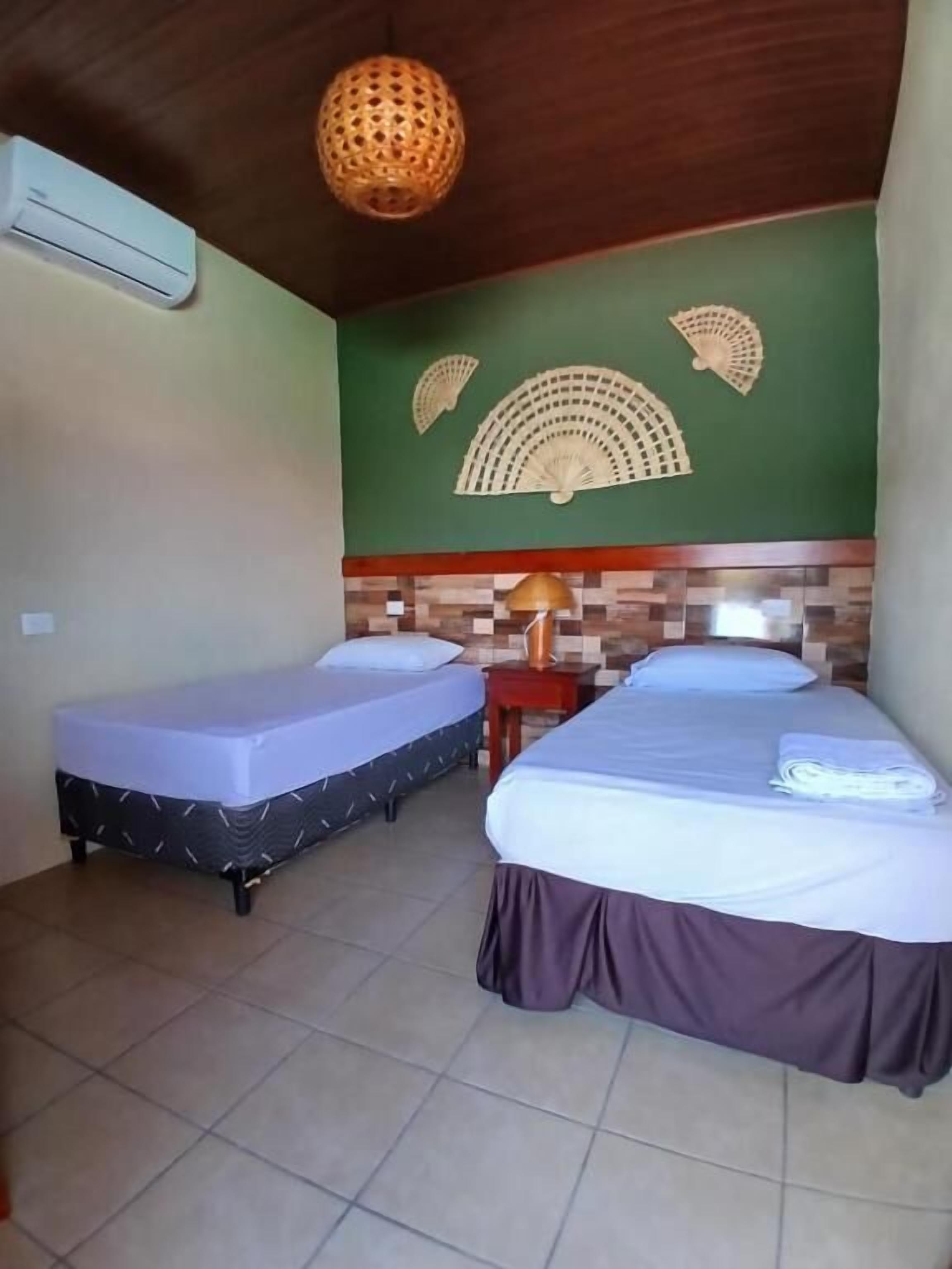 Duas camas de solteiro com cabeceiras, um ventilador de parede, um abajur e piso de azulejos.