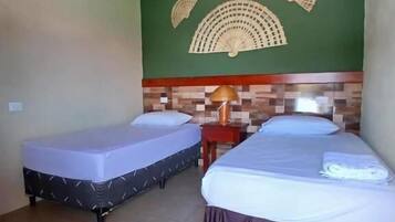 Duas camas de solteiro com cabeceiras, um ventilador de parede, um abajur e piso de azulejos.
