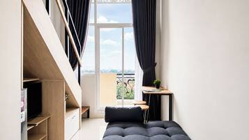 Quarto moderno com cama estilo loft, uma pequena mesa com uma planta e vista para a cidade através de uma grande janela.