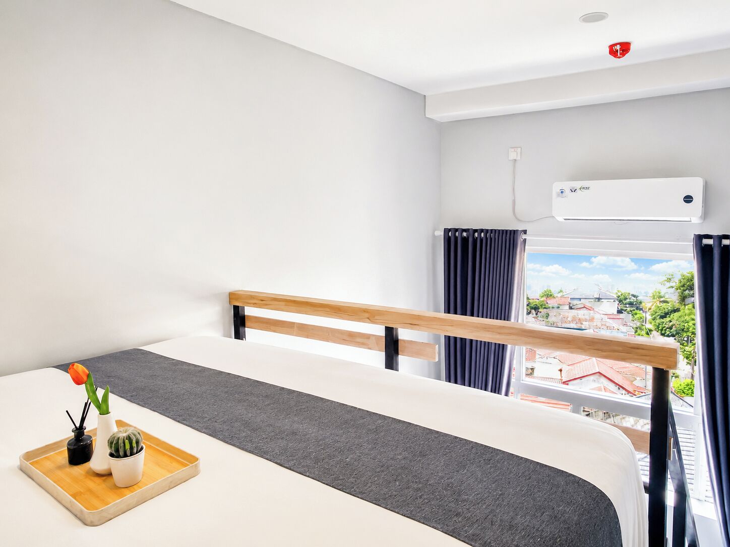 Quarto de hotel com cama, uma pequena mesa com um vaso de flores, uma planta em vaso e uma janela com cortinas.