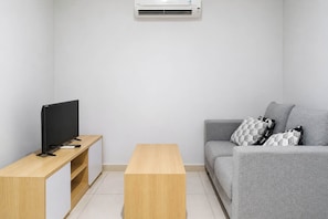 Uma sala de estar minimalista com uma televisão de tela plana em um suporte de madeira, um sofá cinza com almofadas estampadas e uma pequena mesa de madeira.