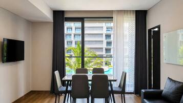 Quarto de hotel moderno com mesa de jantar, quatro cadeiras, televisão de tela plana, sofá de couro preto e uma grande janela com vista para um edifício e áreas verdes.