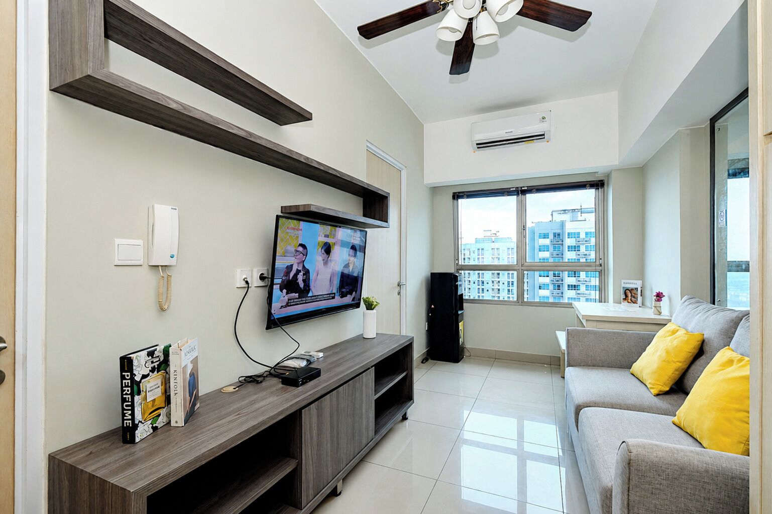 Uma sala de estar moderna com televisão de tela plana, mesa de centro, sofá com almofadas amarelas, ventilador de teto e vista para a cidade através de janelas amplas.