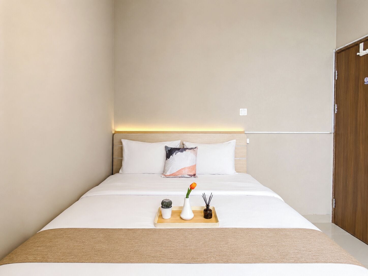 Quarto de hotel com uma cama, uma mesa de cabeceira com bandeja, um vaso com uma flor e um pequeno cacto.