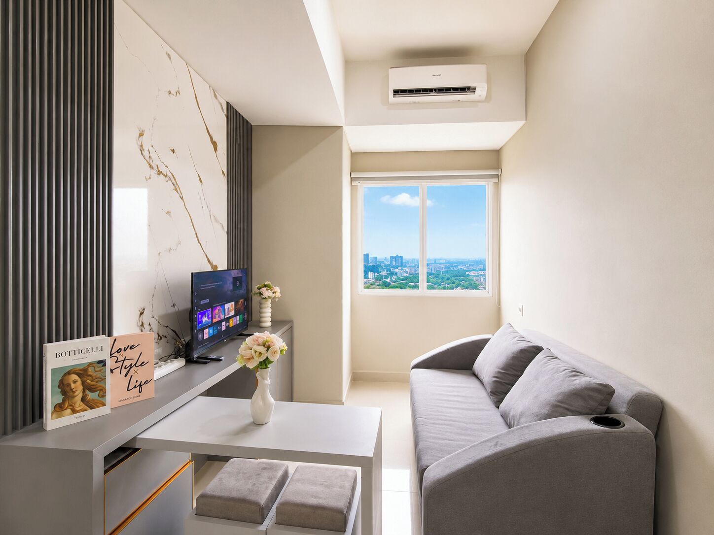 Uma sala de estar moderna com um sofá cinzento, uma televisão de tela plana, uma mesa de centro branca com um vaso de flores e vista para a cidade por uma janela grande.