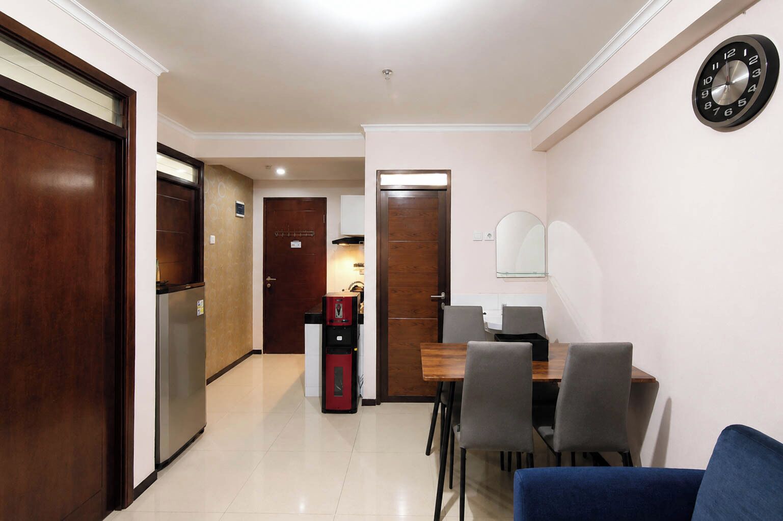 Quarto de hotel com porta de madeira, área de jantar com mesa e cadeiras, máquina de café e um relógio na parede.