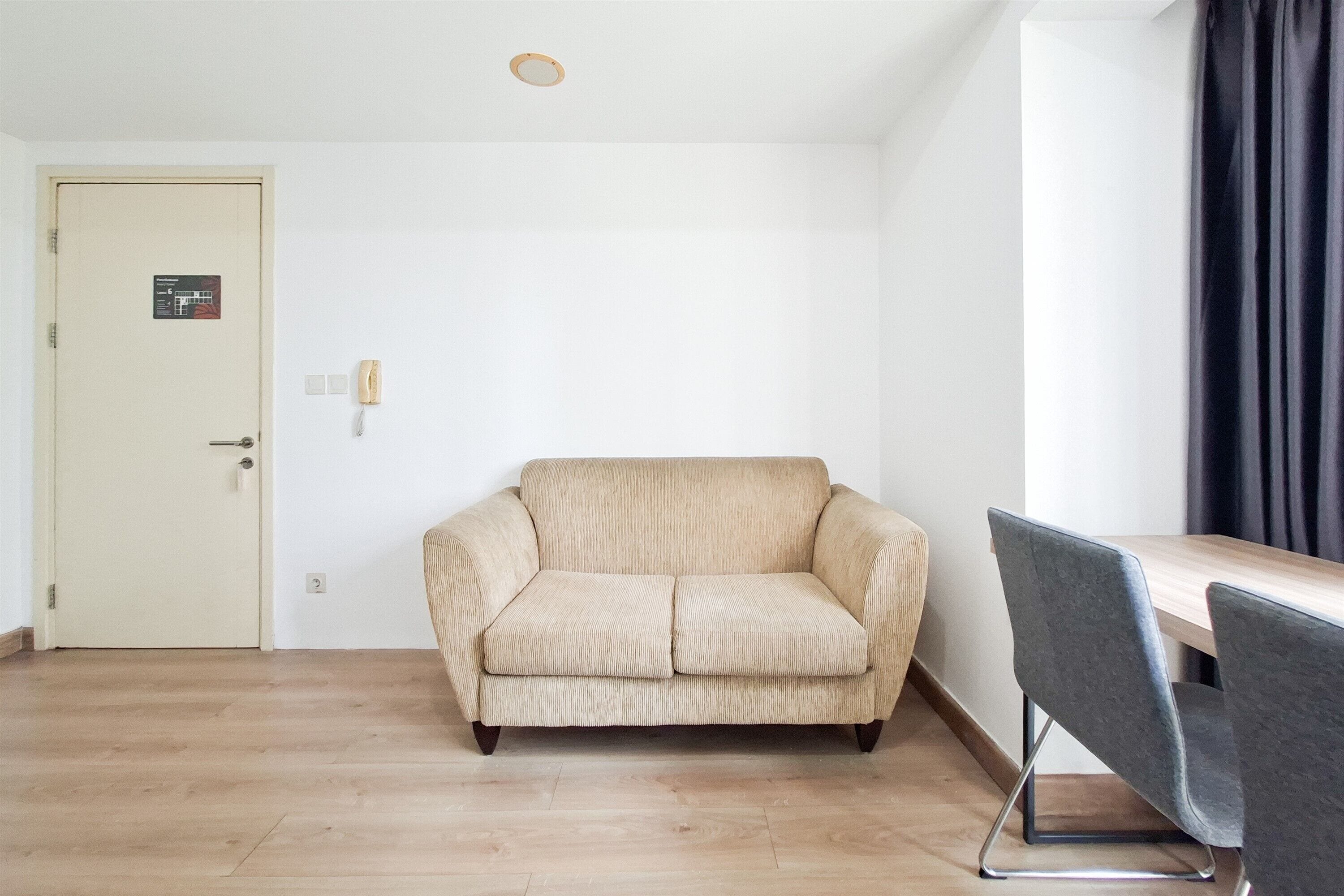 Um quarto com sofá bege, uma mesa de madeira, uma cadeira, uma porta com um letreiro e um lustre no teto.