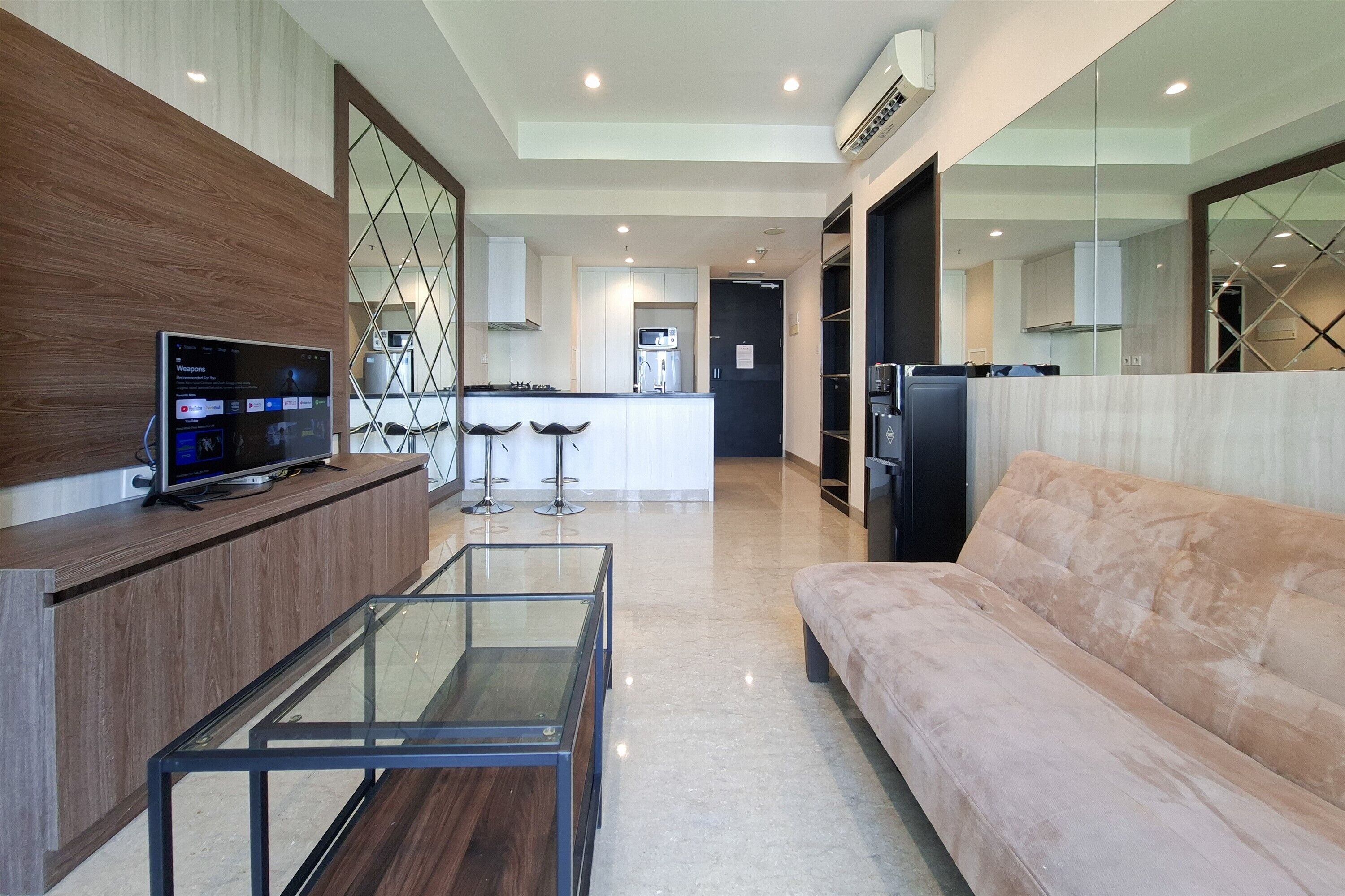 Uma sala de estar moderna com uma mesa de centro de vidro, um sofá, uma TV sobre um móvel de madeira e uma cozinha ao fundo.