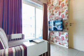 Quarto de hotel com cama, escrivaninha com teclado, televisão na parede e janela com cortinas.