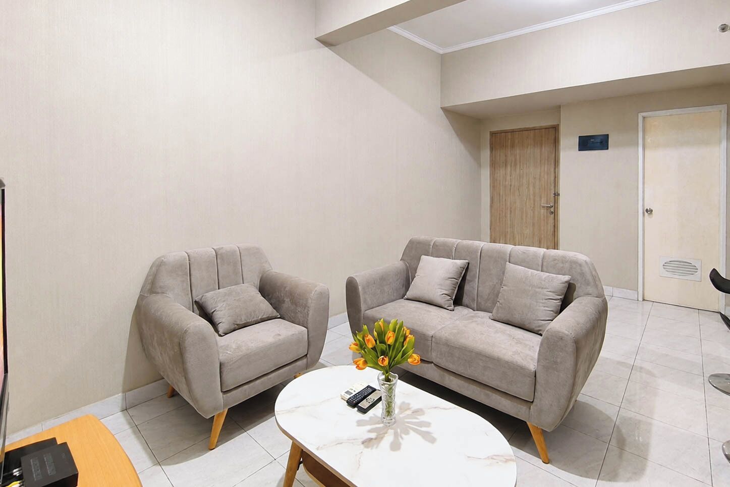 Quarto moderno com dois sofás cinzentos, uma pequena mesa branca com um vaso de flores e uma porta ao fundo.