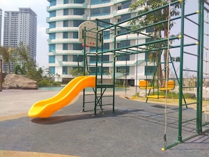 Um parquinho com um escorrega amarelo e estruturas de escalada, cercado por um edifício com vários andares e janelas.