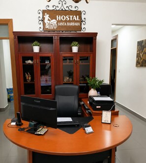 Uma mesa de recepção com um monitor Planar, uma cadeira de escritório preta, um armário de madeira com portas de vidro e uma placa com a inscrição 'HOSTAL SANTA BARBARA'.