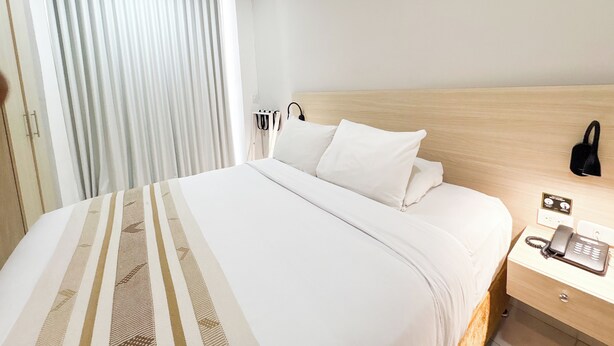 Quarto de hotel com cama, mesa de cabeceira com telefone e janela com cortinas.