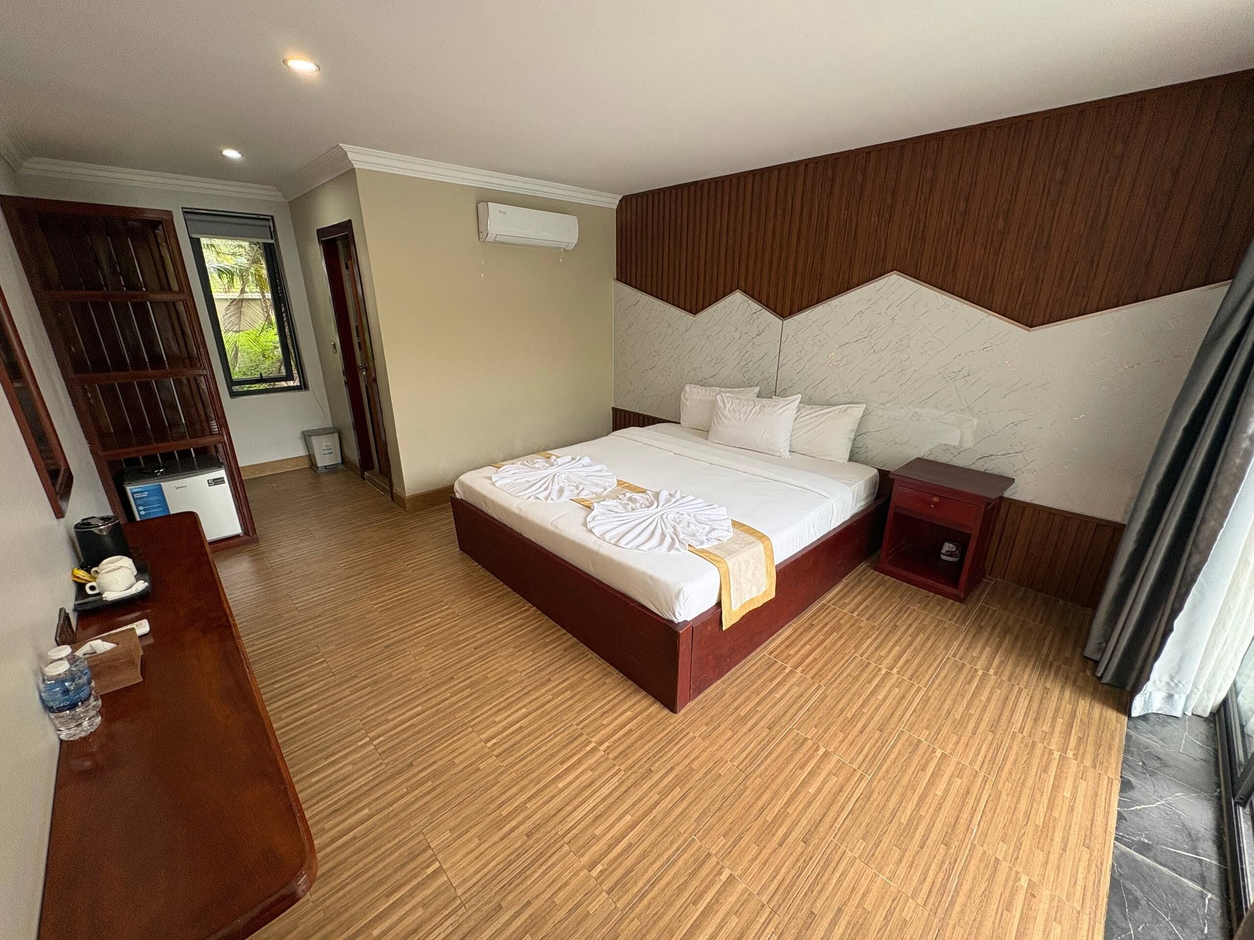 Quarto de hotel com cama grande, piso de madeira, escrivaninha, mesa de cabeceira e janela com vista.