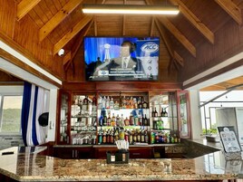 Uma área de bar com uma televisão exibindo o início do campeonato de futebol ACC, prateleiras repletas de diversas garrafas e um balcão com um painel de menu.