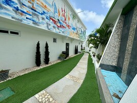 Corredor de hotel com grama artificial, um caminho de pedra, um mural na parede e área de piscina.