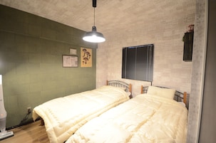 Quarto com duas camas, uma luminária no teto, janela com persianas e prateleira fixada na parede.