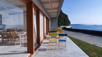 Uma área de refeições moderna ao ar livre com vista para o mar, cadeiras de madeira e um deck de concreto.