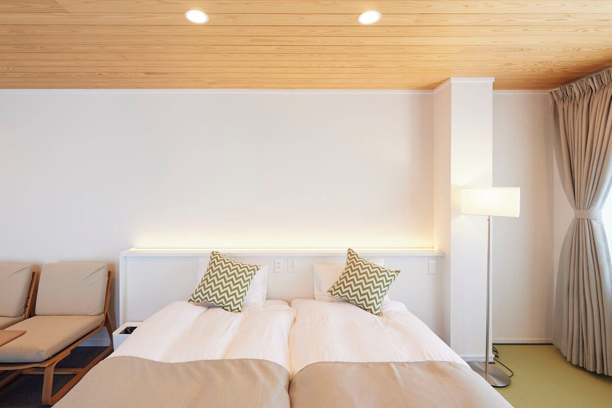 Quarto de hotel com uma cama, duas cadeiras, uma mesa de cabeceira, uma lâmpada e uma janela com cortinas.