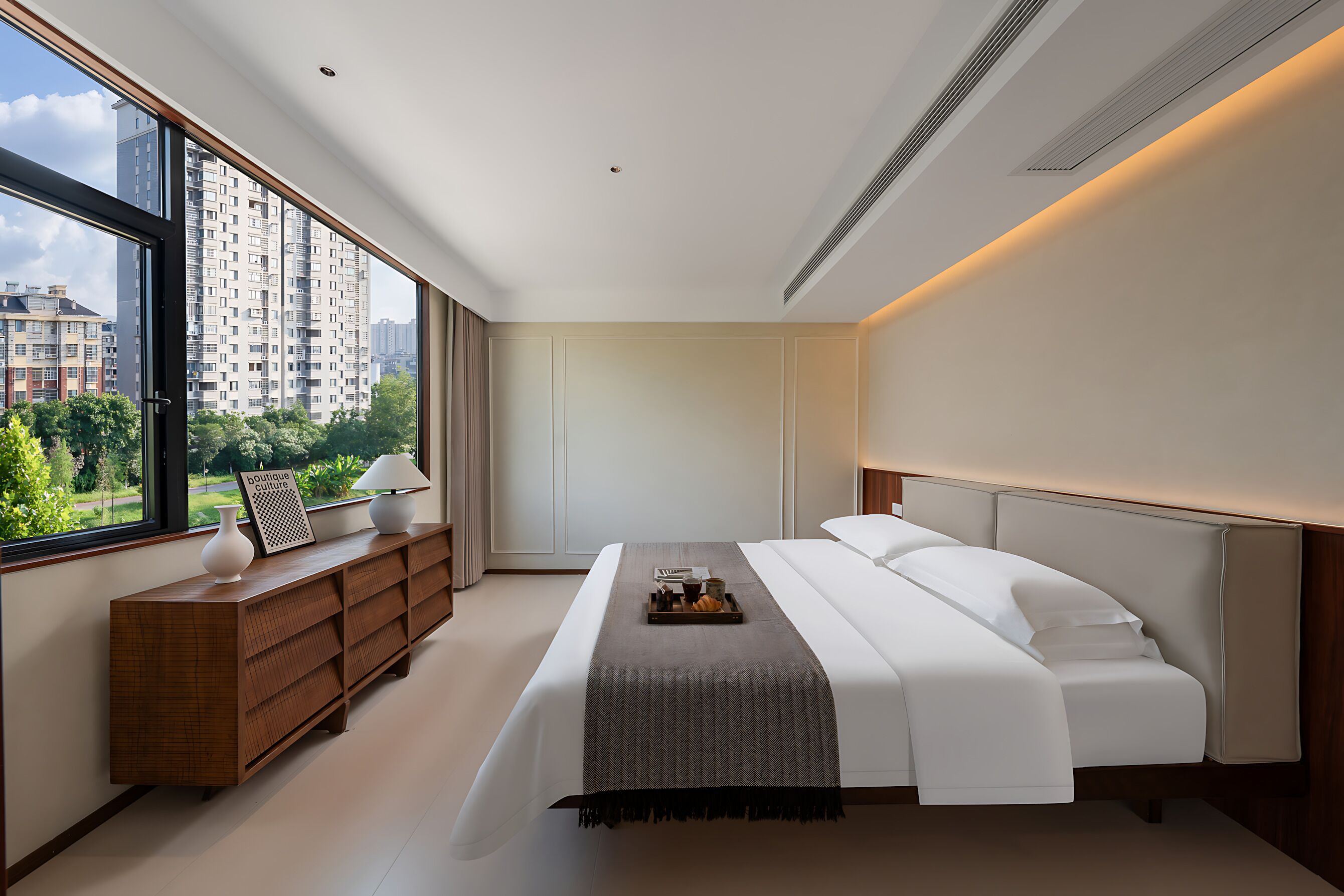 Quarto de hotel moderno com uma janela grande, uma cama com roupa de cama branca, uma mesa consola de madeira e vista para a paisagem urbana.
