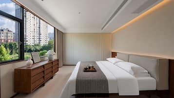 Quarto de hotel moderno com uma janela grande, uma cama com roupa de cama branca, uma mesa consola de madeira e vista para a paisagem urbana.