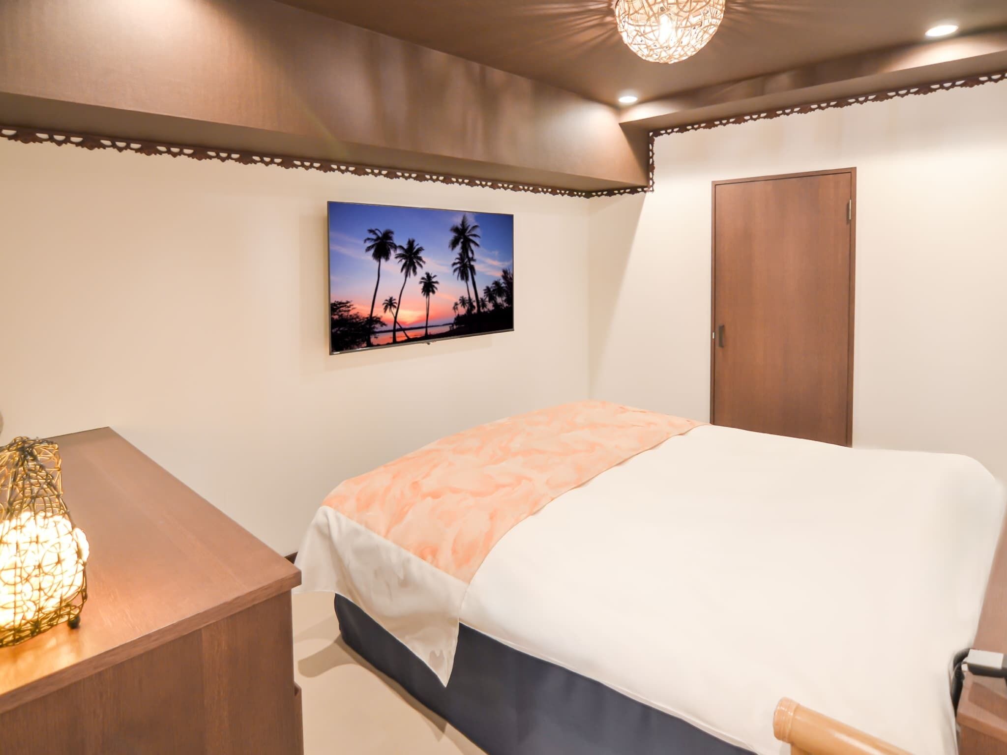 Quarto com cama, escrivaninha, cadeira, porta e uma imagem pendurada na parede de um pôr do sol com palmeiras.