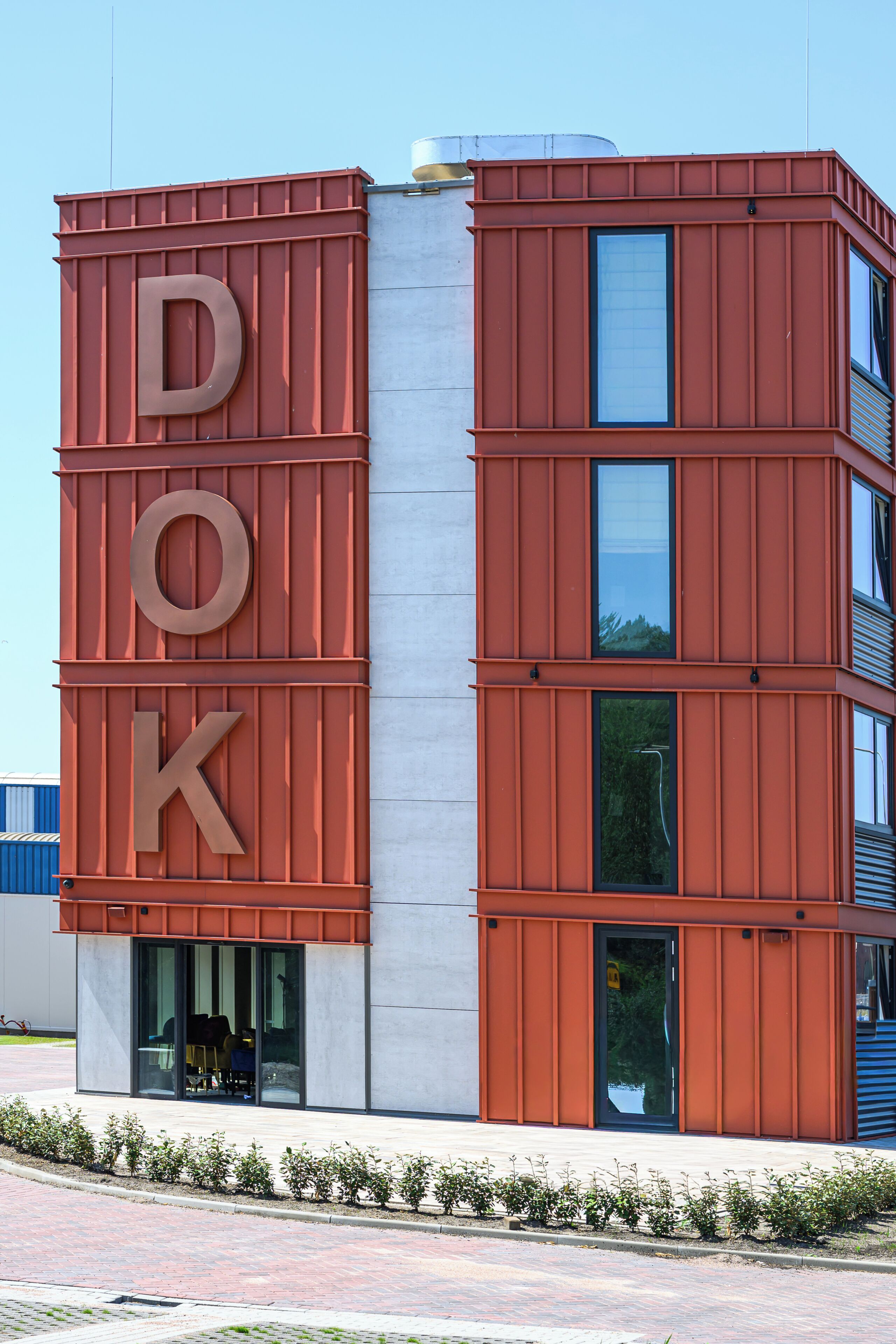 Um edifício moderno com um letreiro 'DOK' em destaque, apresentando uma estrutura com vários andares, grandes janelas e telhado plano.